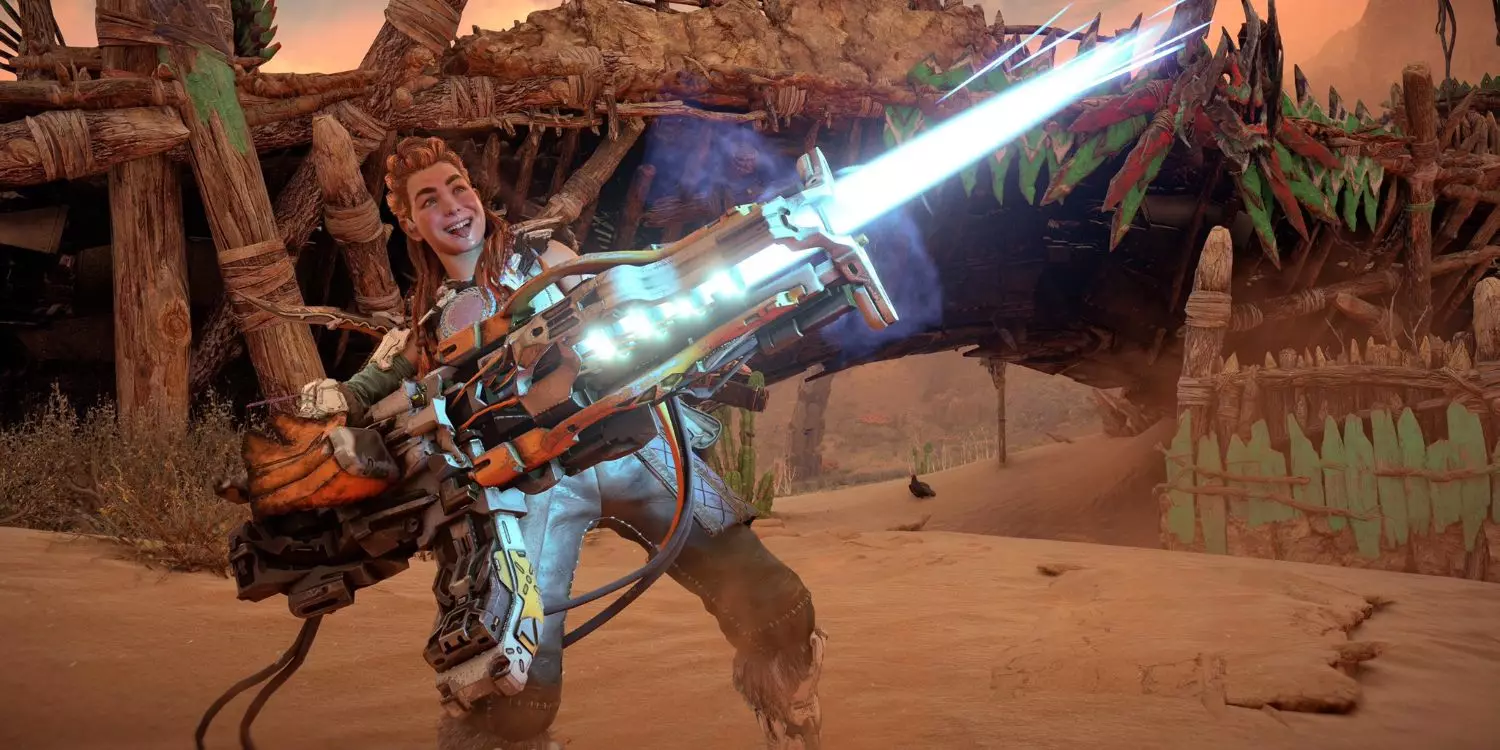 aloy, dengan seringai di wajahnya dan lidahnya menjulur, memegang pistol besar dengan sinar biru bercahaya yang keluar dari nozzle