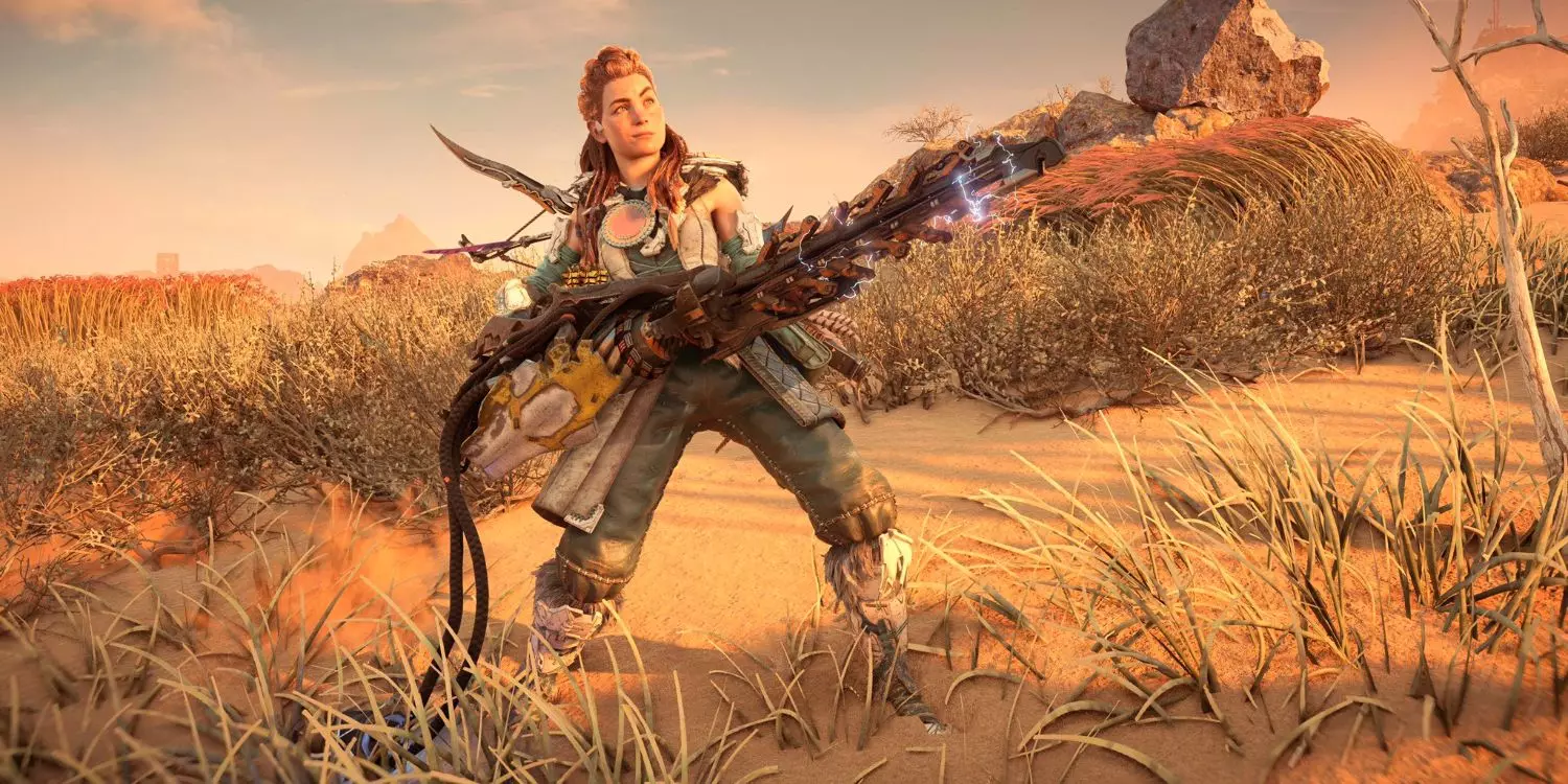 aloy memegang pistol besar dengan laras panjang yang menyala sambil berdiri di padang pasir dengan rumput pendek dan alang-alang di dekatnya