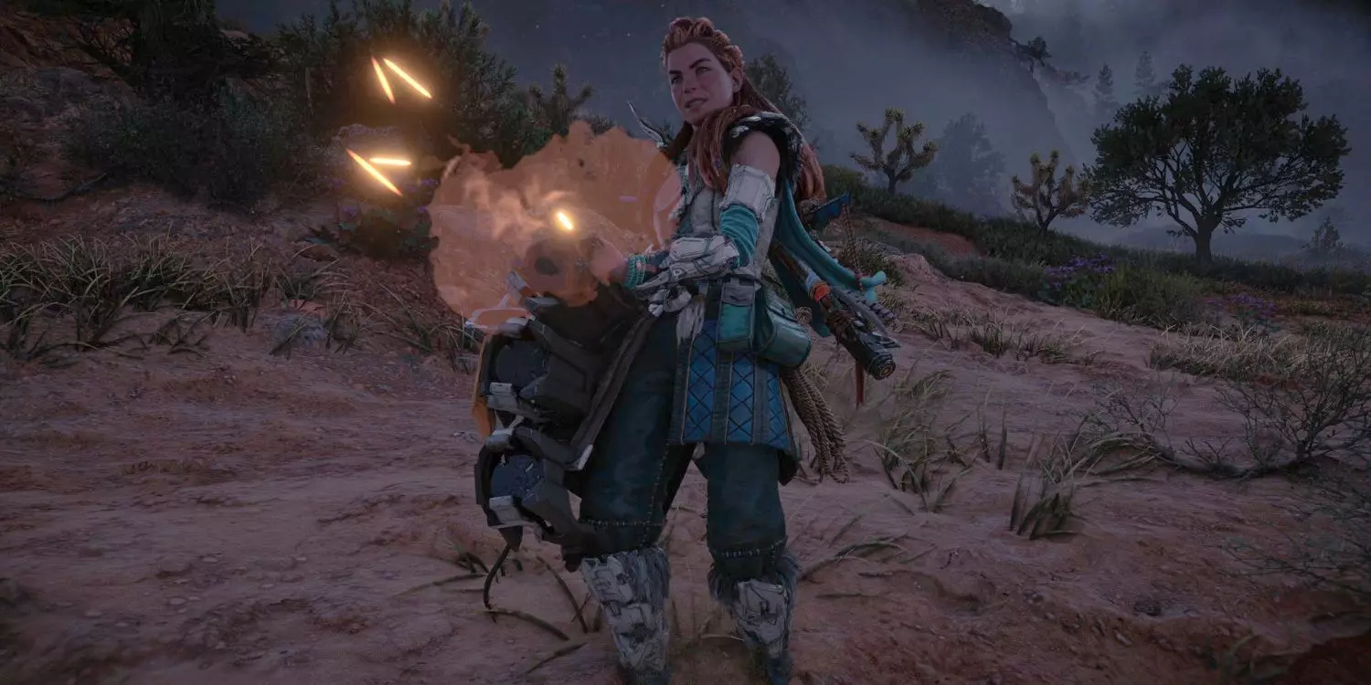 Η Aloy, με μια πράσινη και λευκή στολή με μεταλλικές πλάκες πάνω από τις κνήμες και τους πήχεις της, κρατά ένα βαρύ κομμάτι μηχανήματος που έχει ένα σύννεφο καπνού και αιωρούμενες χόβολες γύρω από το ακροφύσιο